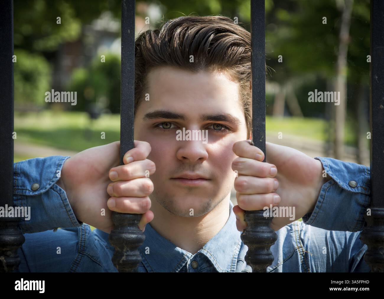 Attractive young man behind metal gate, gripping bars with his hands ...
