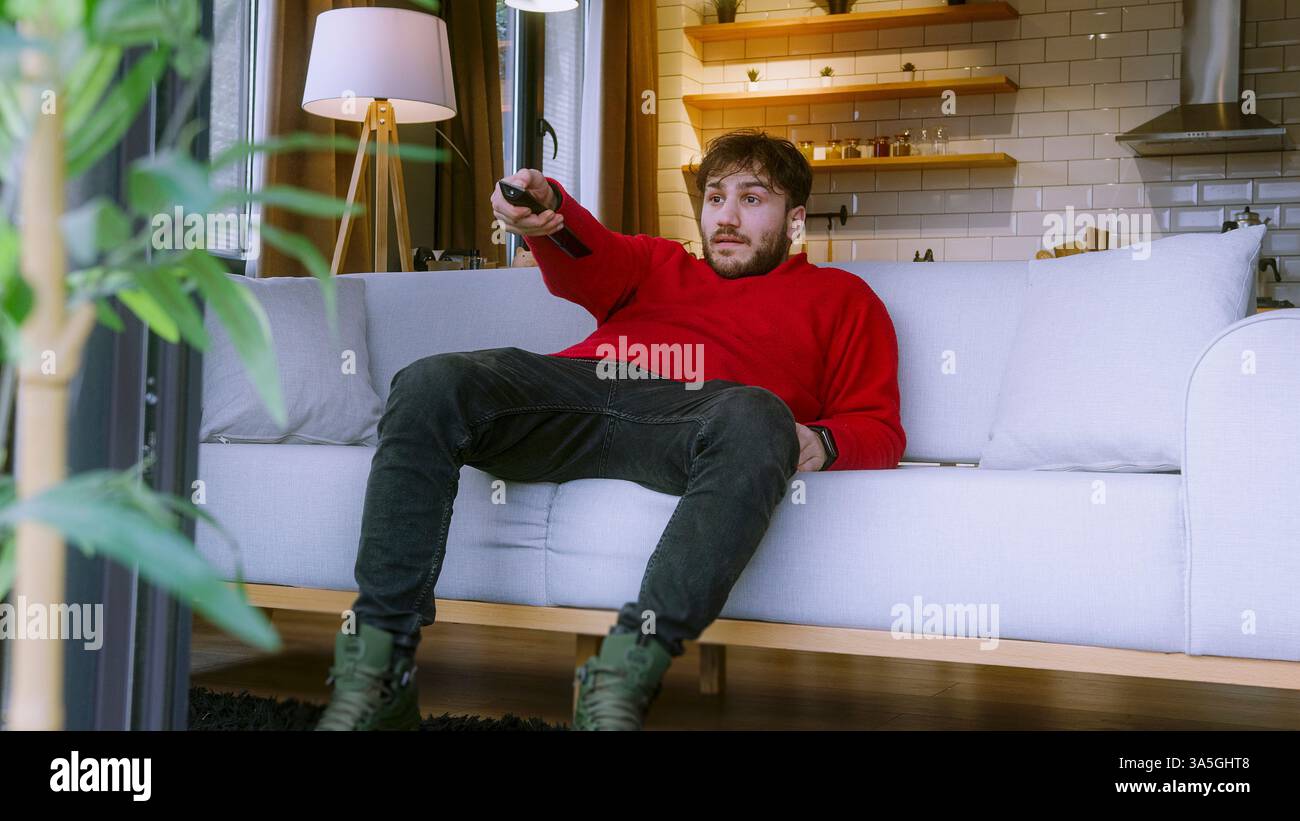 Relaxed Young Man Watching TV at Home, Sitting Comfortably on the Couch with a Remote Control in ...