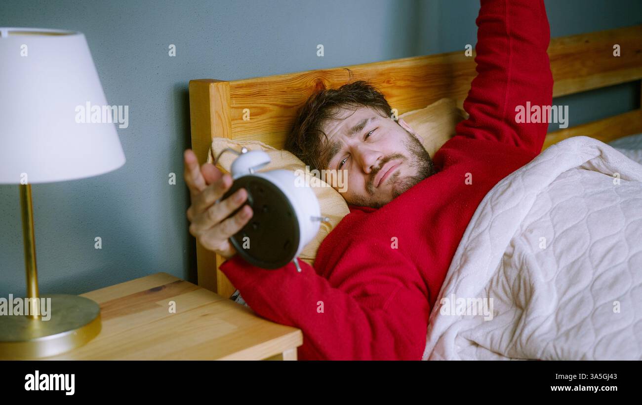 Young Sleepy Man Waking Up and Yawning in Bed with Alarm Clock Ringing ...