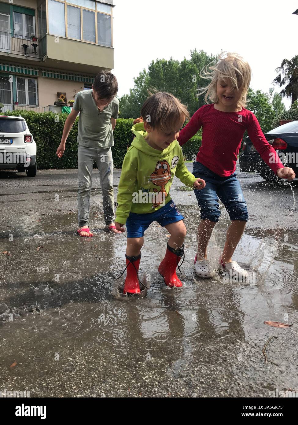 Three children joyfully splash in a large puddle, wearing colorful ...