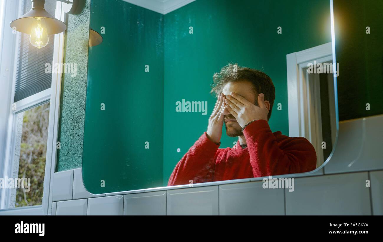 Man in a red sweater standing in front of a mirror, covering his ears ...