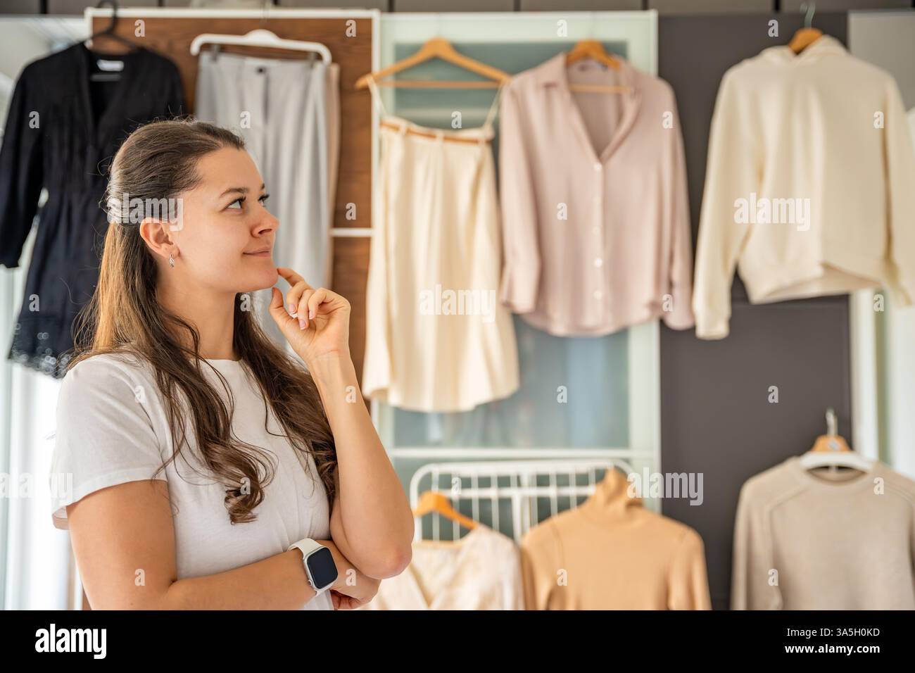 A young woman stands in front of a wardrobe filled with hanging outfits ...