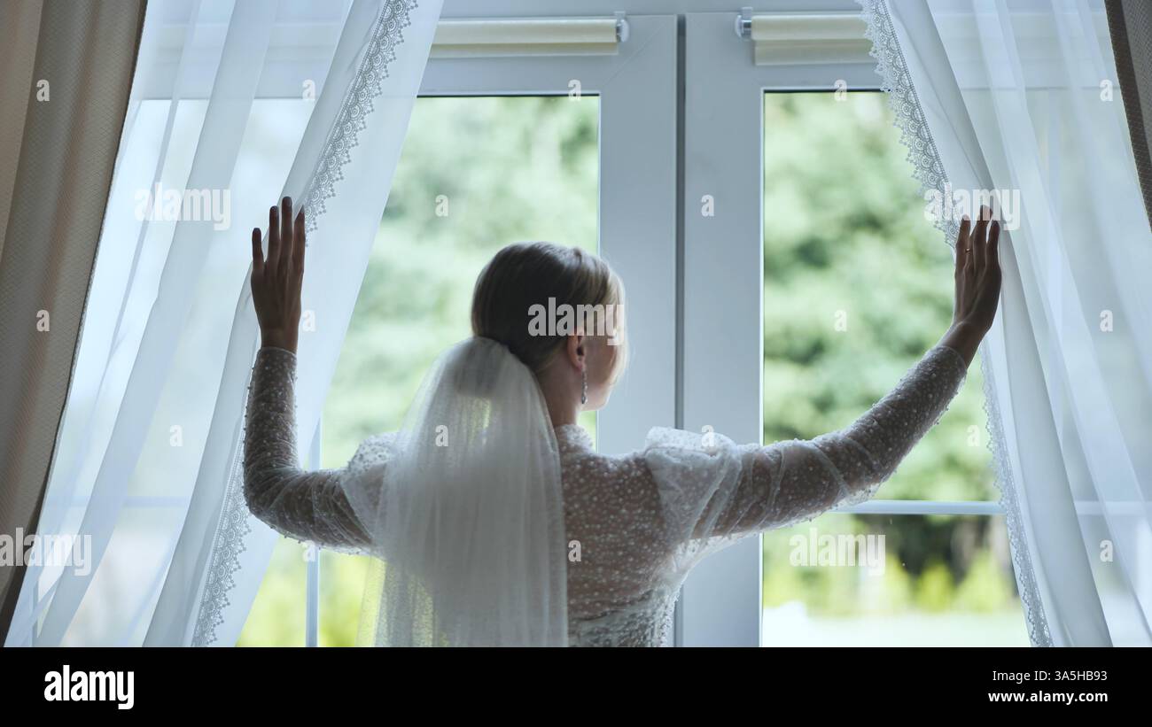 White gowned bride pulling back curtains, gazing through window at ...