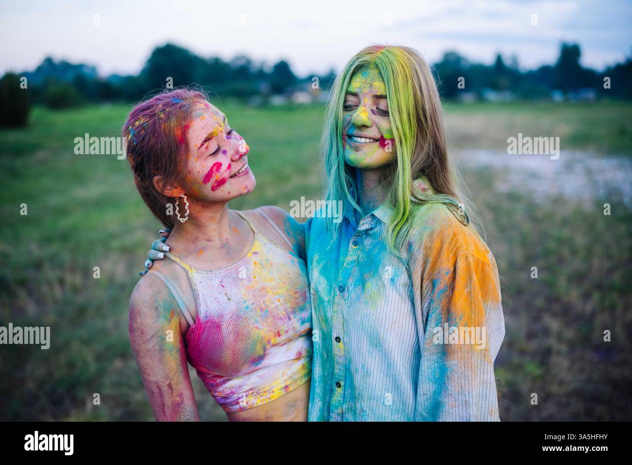 Two young women, covered in vibrant holi powder, embrace and smile at a ...