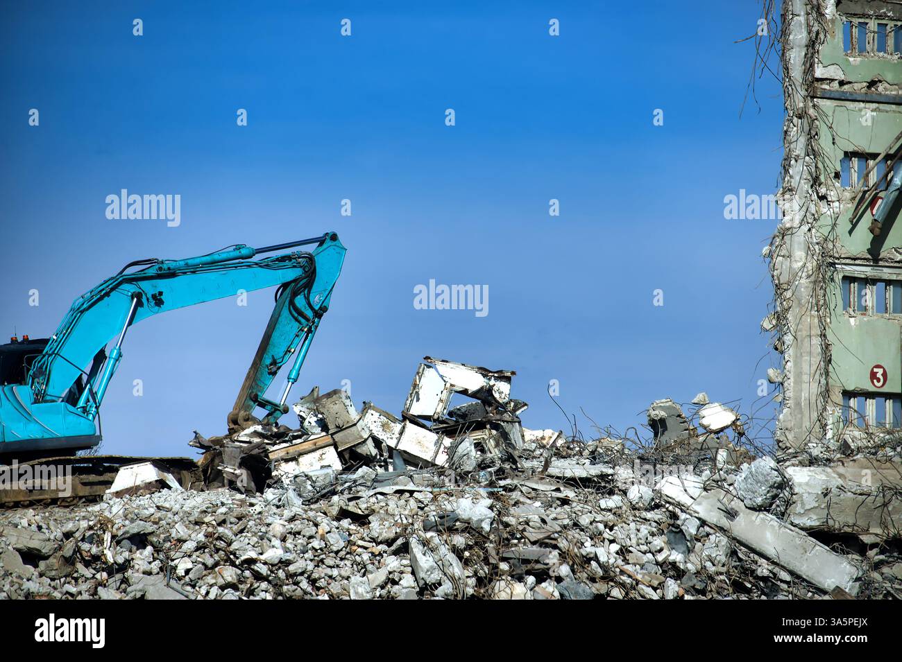 A teal excavator stands atop a pile of rubble with a blue sky ...
