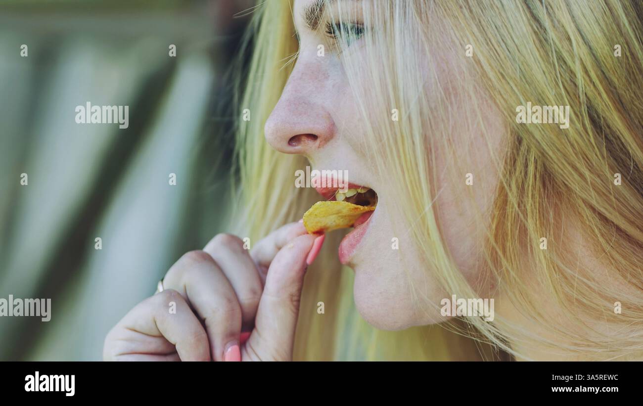 Blond woman biting crunchy potato chip, smiling while tasting salty ...