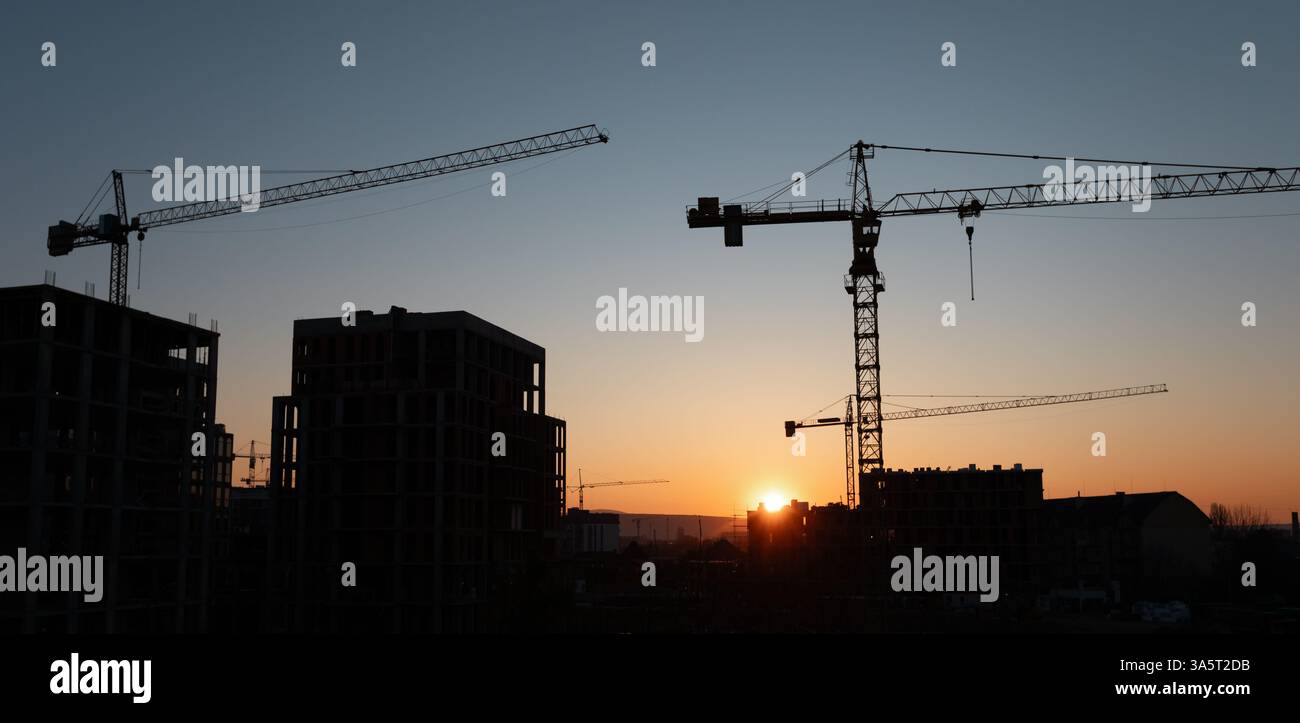 Construction cranes tower over a developing urban area as the sun sets ...
