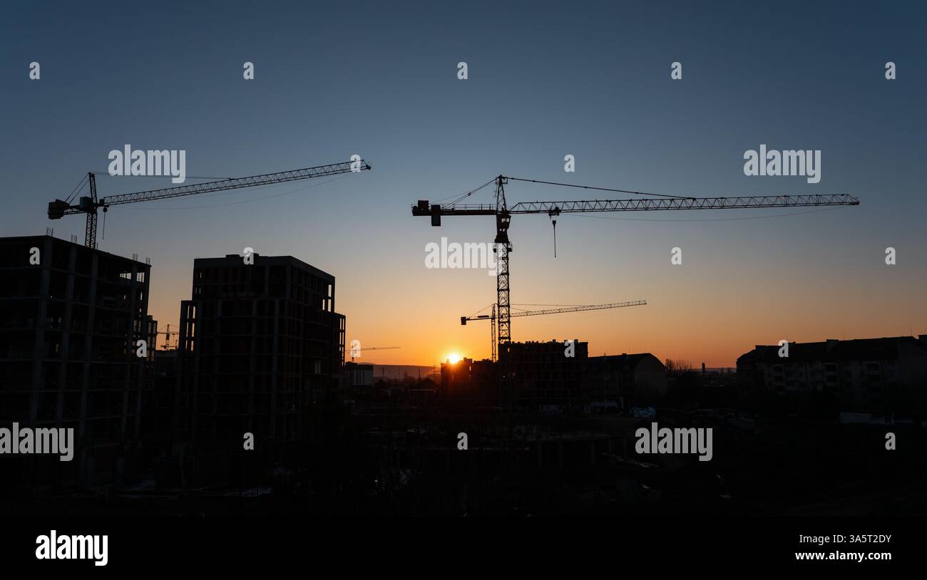 Cranes tower over a developing urban landscape at sunset, casting long ...