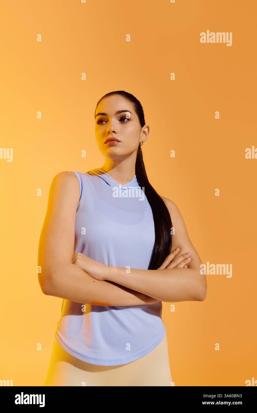 A confident young woman with arms crossed poses against a warm orange ...