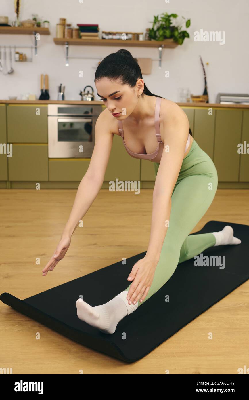 A young woman stretches on a yoga mat in her kitchen, highlighting her ...