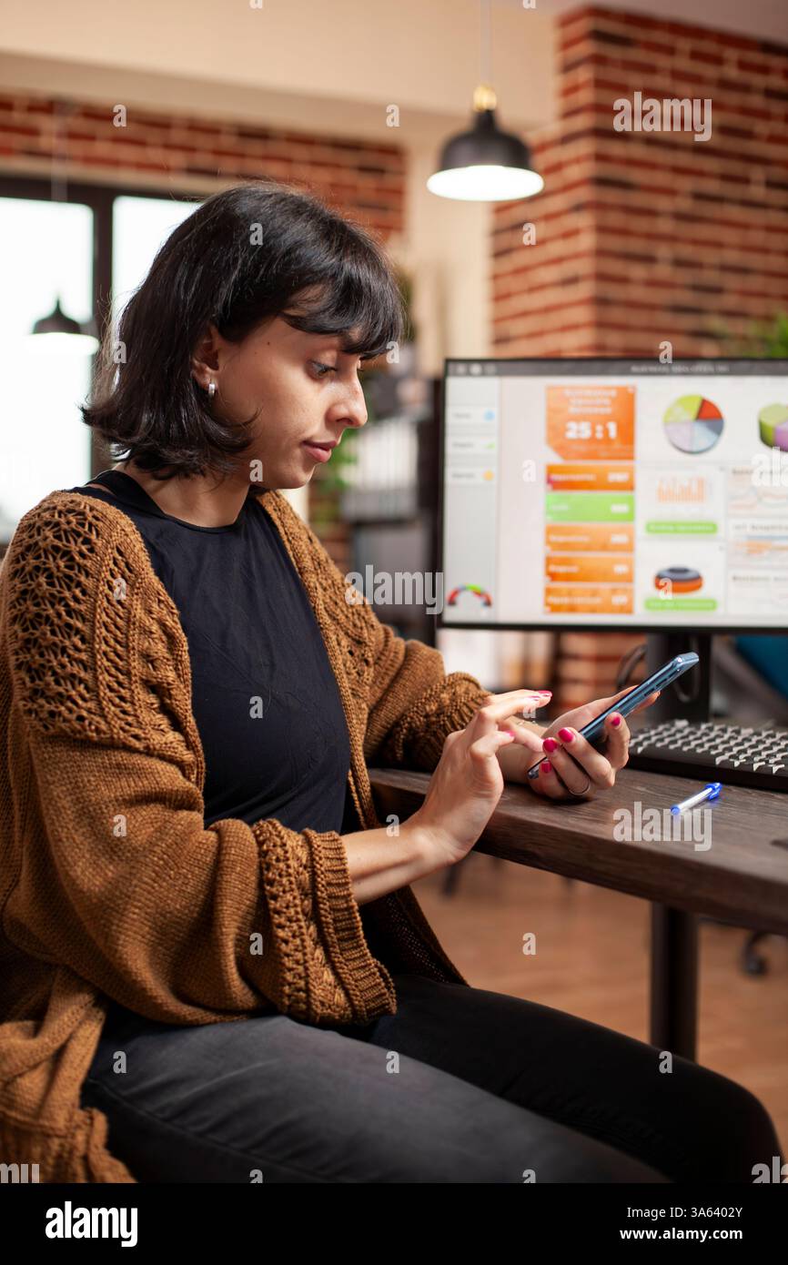 Businesswoman checking emails on smartphone as desktop monitor displays ...