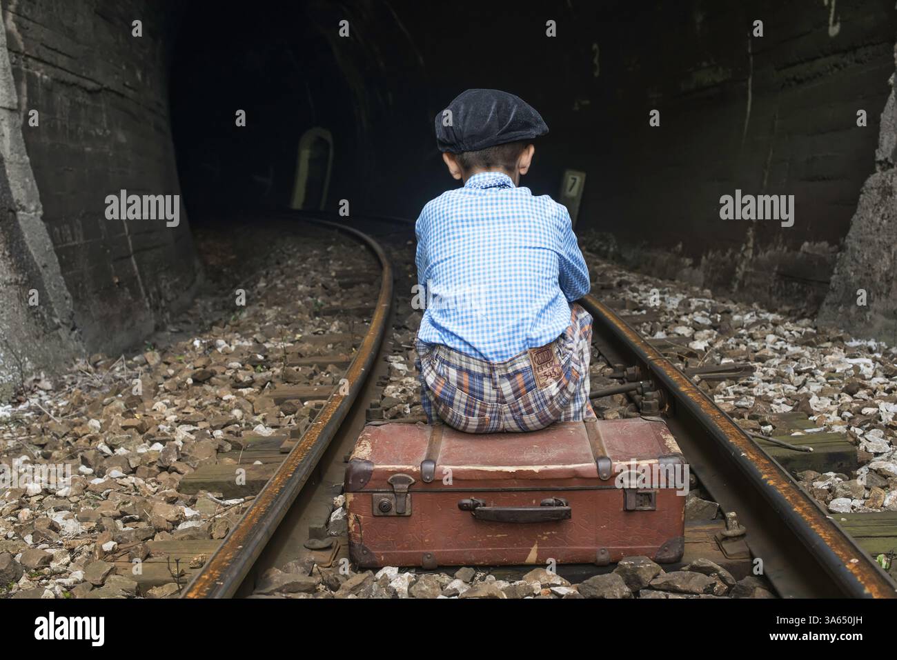 Child in vintage clothes sits on railway road in front of a tunnel ...