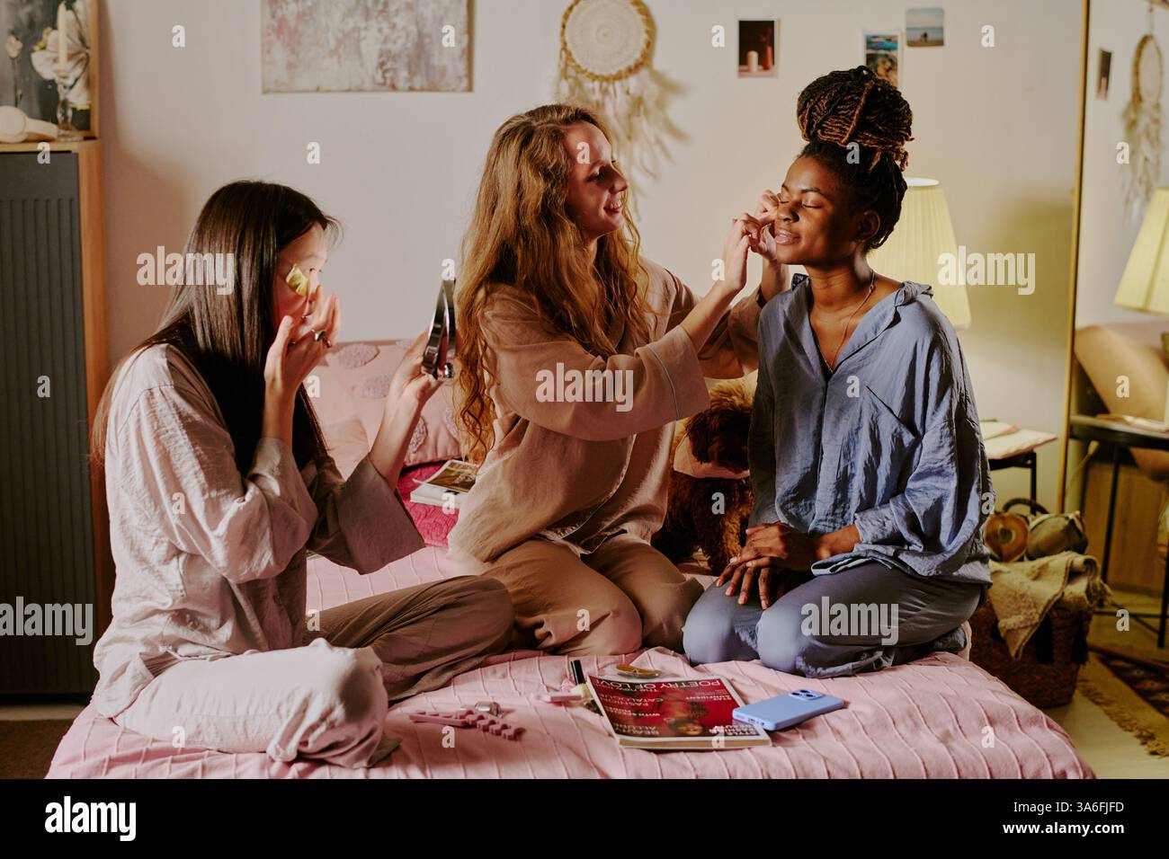 Wide shot of three biracial friends in pajamas sitting on bed and doing ...