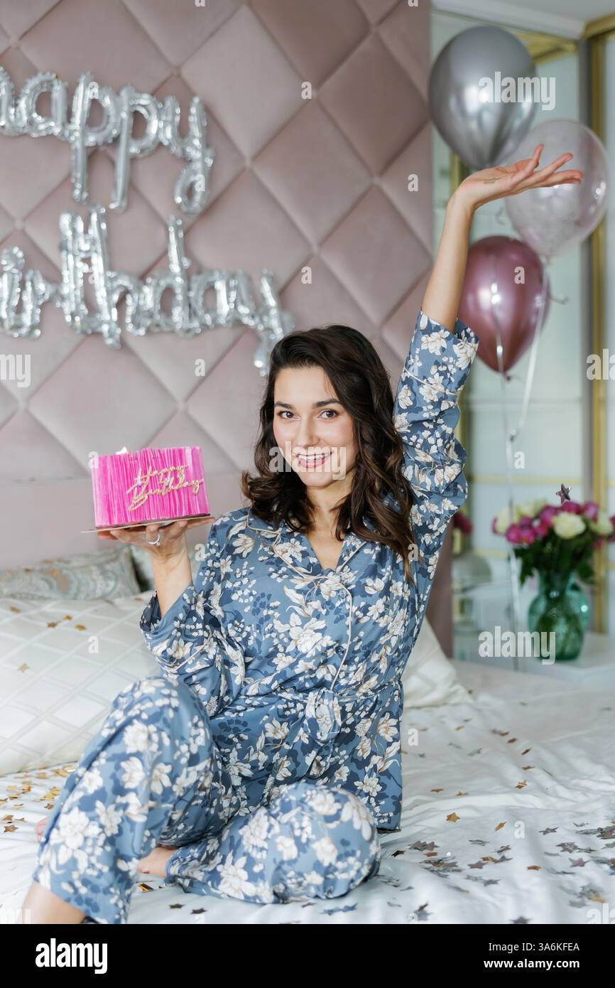 Young woman sitting on bed holding birthday cake and raising arm ...