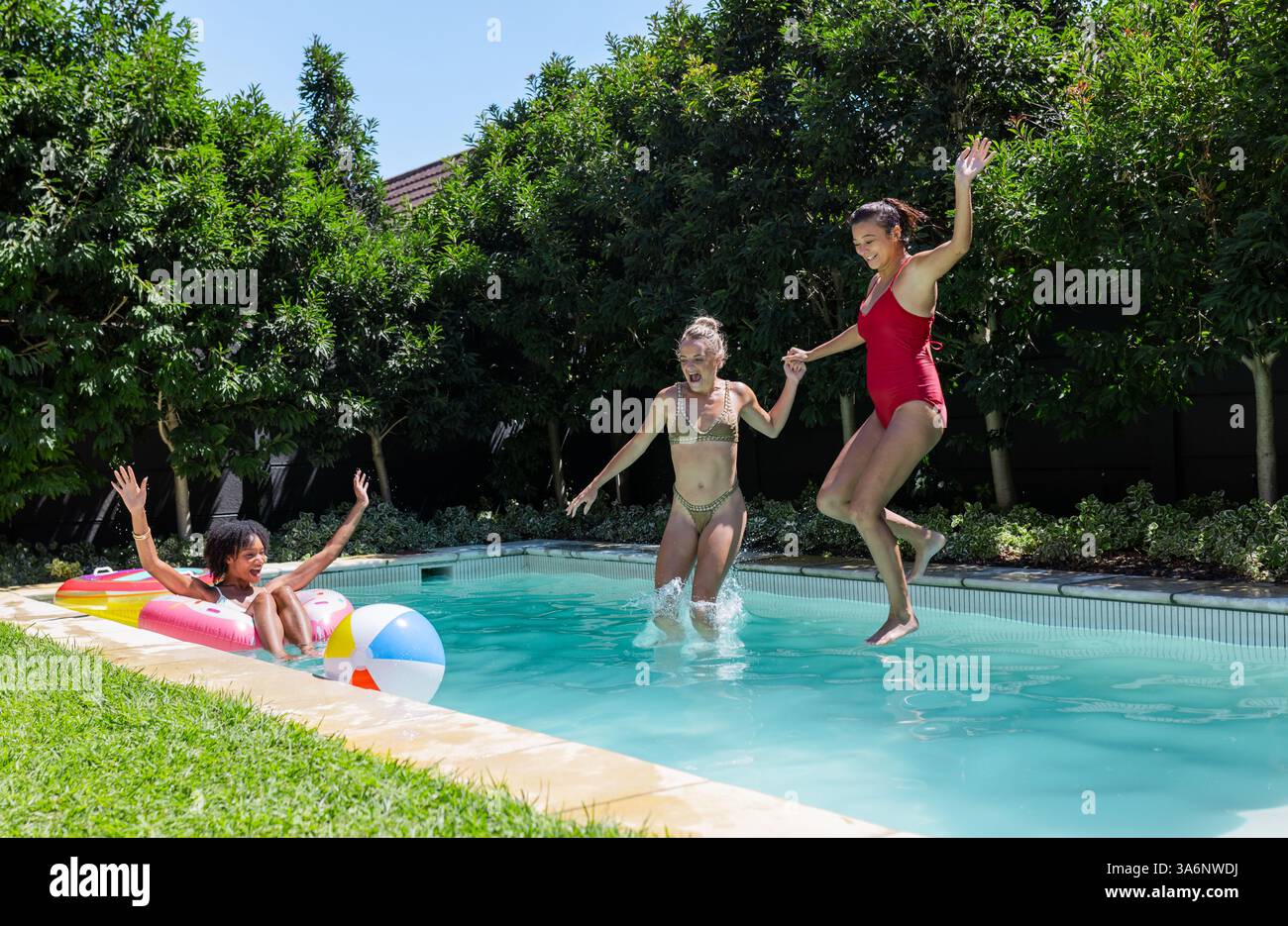 Diverse female friends jumping into pool while another relaxes on float ...