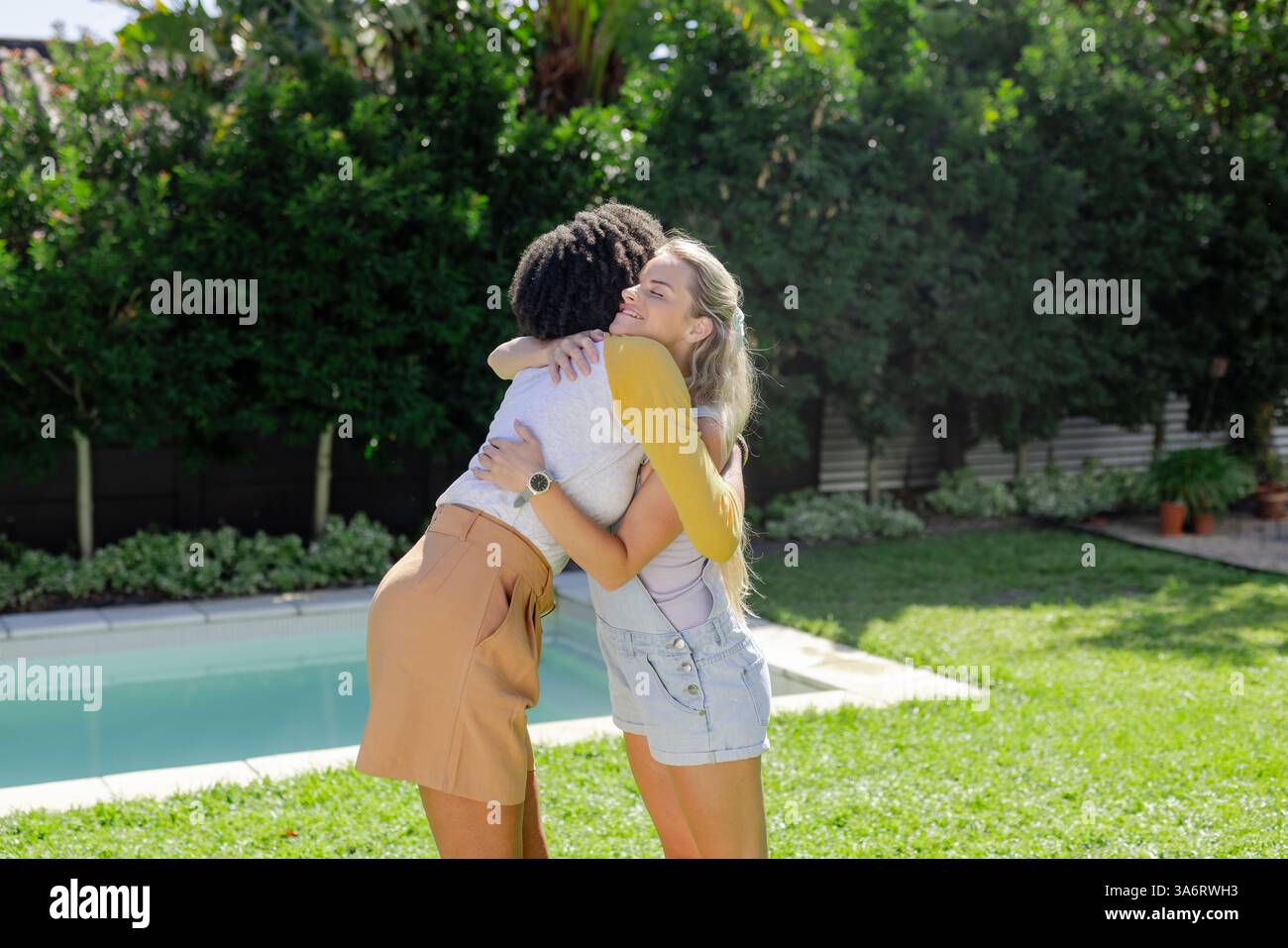 Female friends hugging joyfully by poolside in sunny backyard garden ...