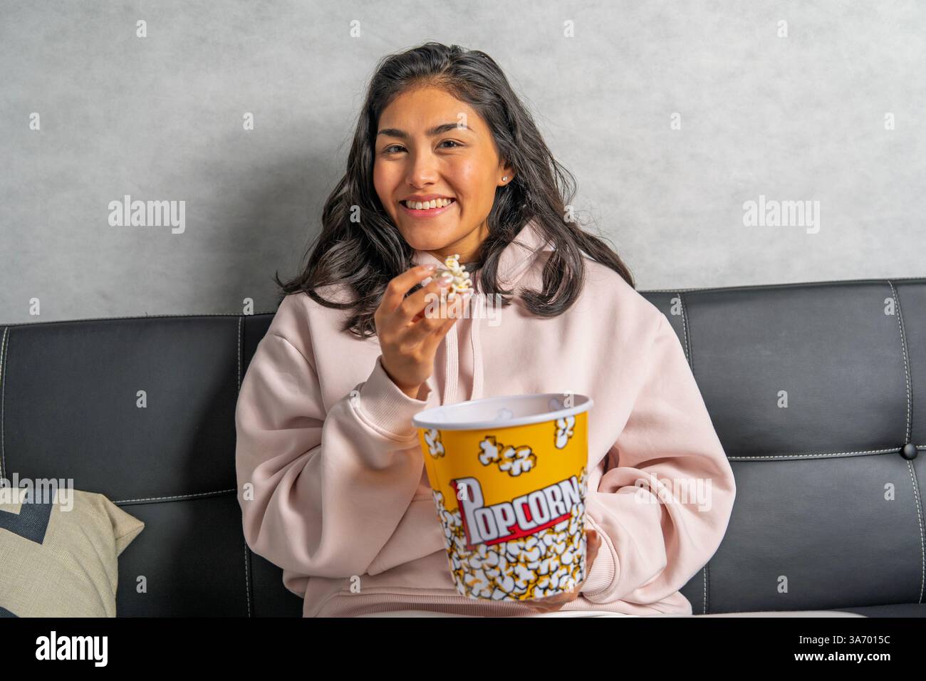Happy young woman sitting on sofa eating popcorn from bucket while ...