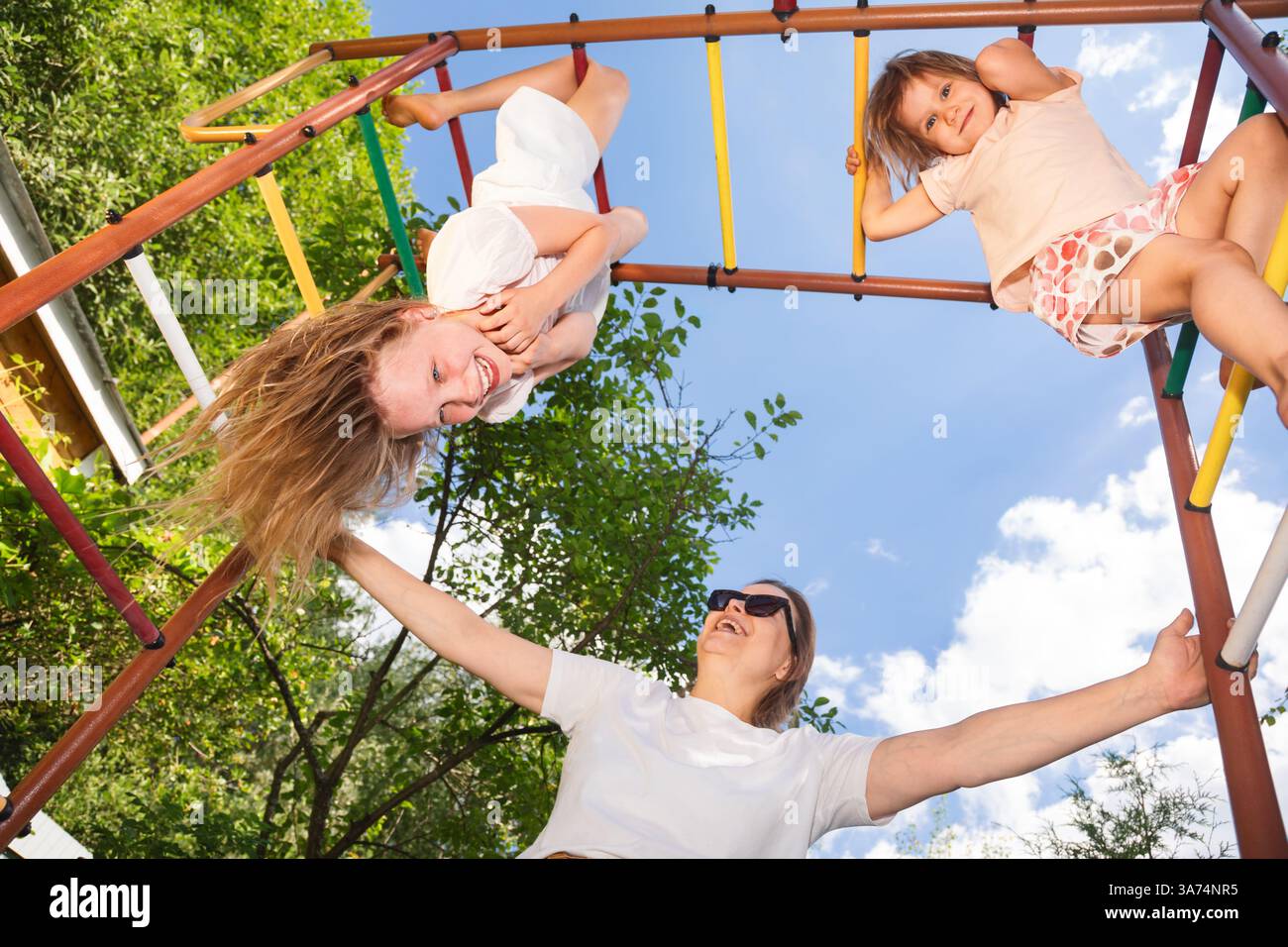 A cheerful mother and her two daughters are playing on a colorful jungle gym in a backyard, with ...