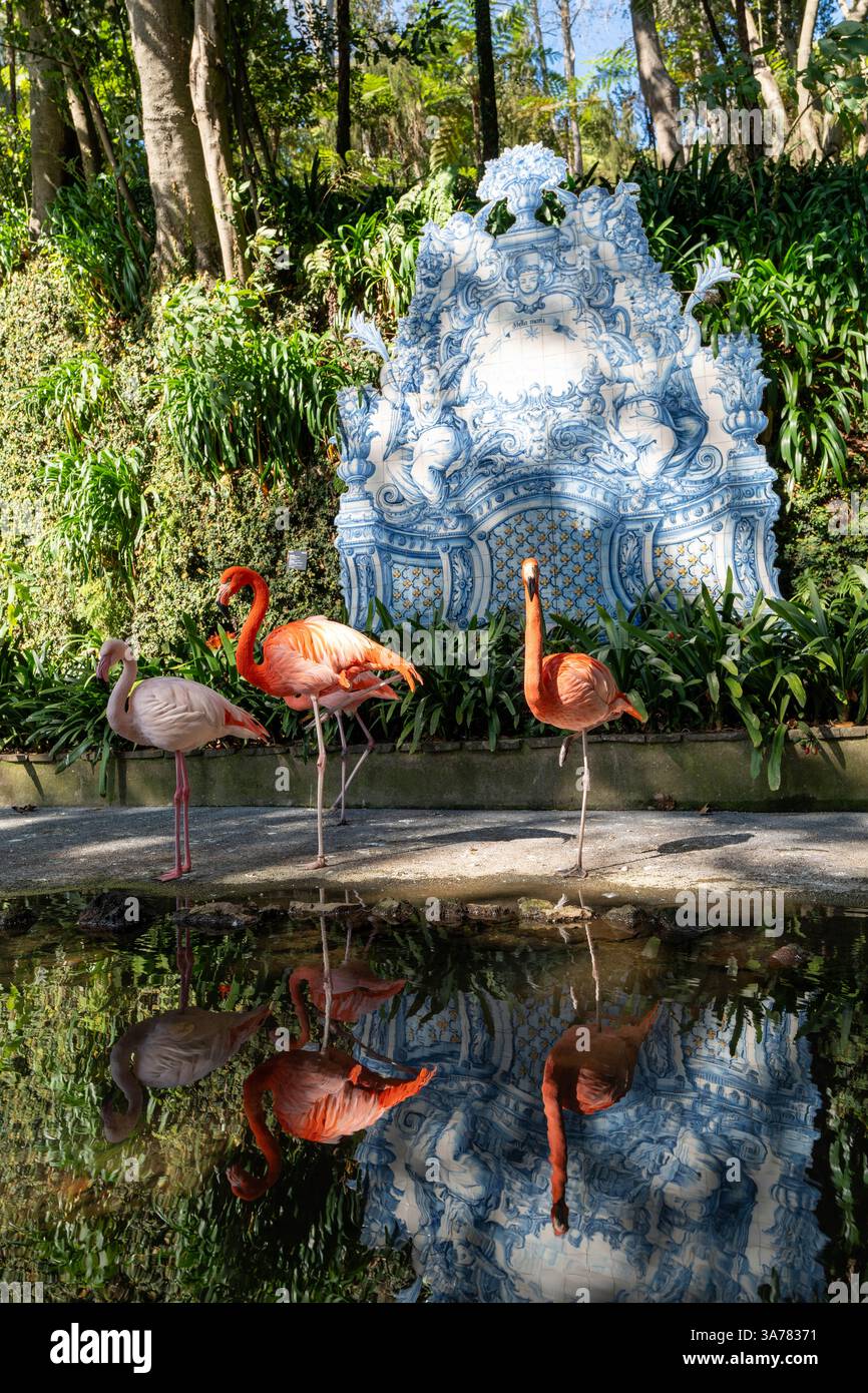 Three pink flamingos stand near a reflective pond in a lush garden. A ...
