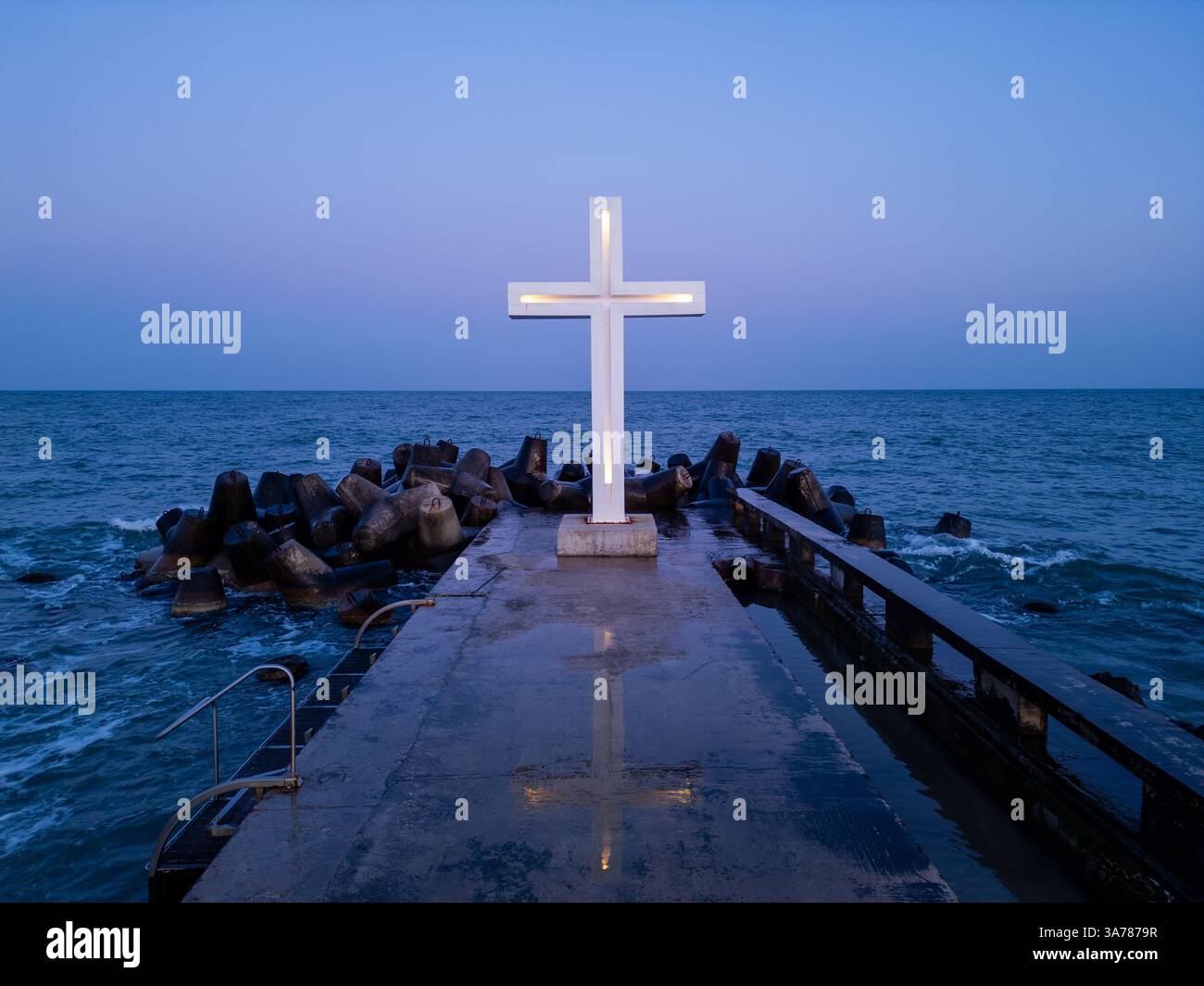 Illuminated cross monument on a concrete pier by the sea at dusk, with ...
