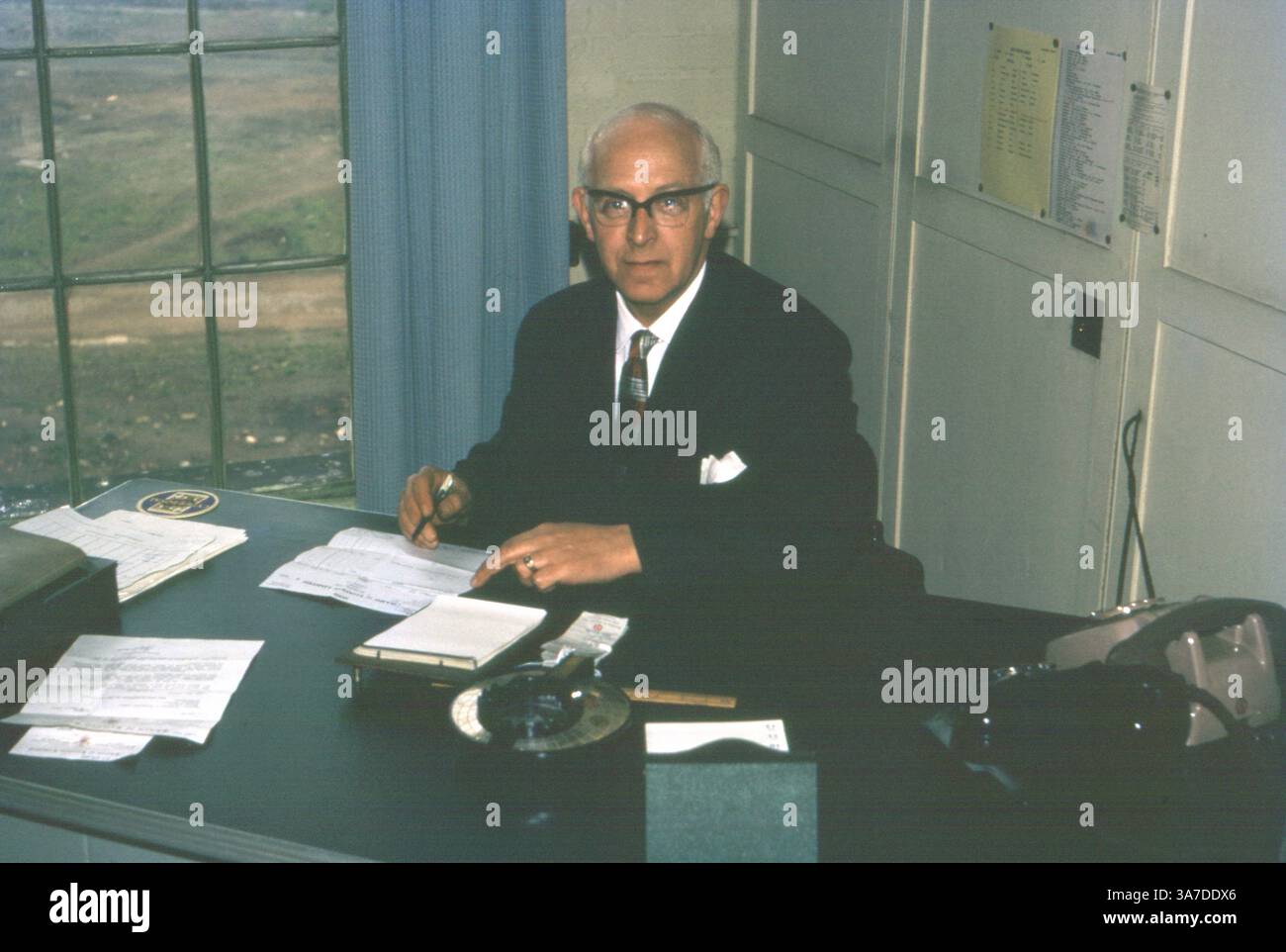 An older man sits at a neatly arranged office desk, pen in hand ...