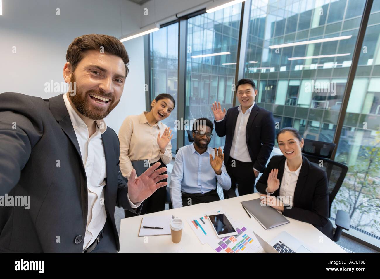 A diverse group of colleagues in a modern office wave to the camera ...
