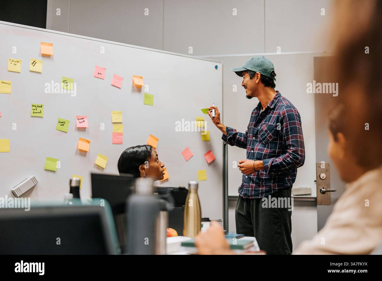 Smiling young man sticking adhesive note on whiteboard while discussing ...