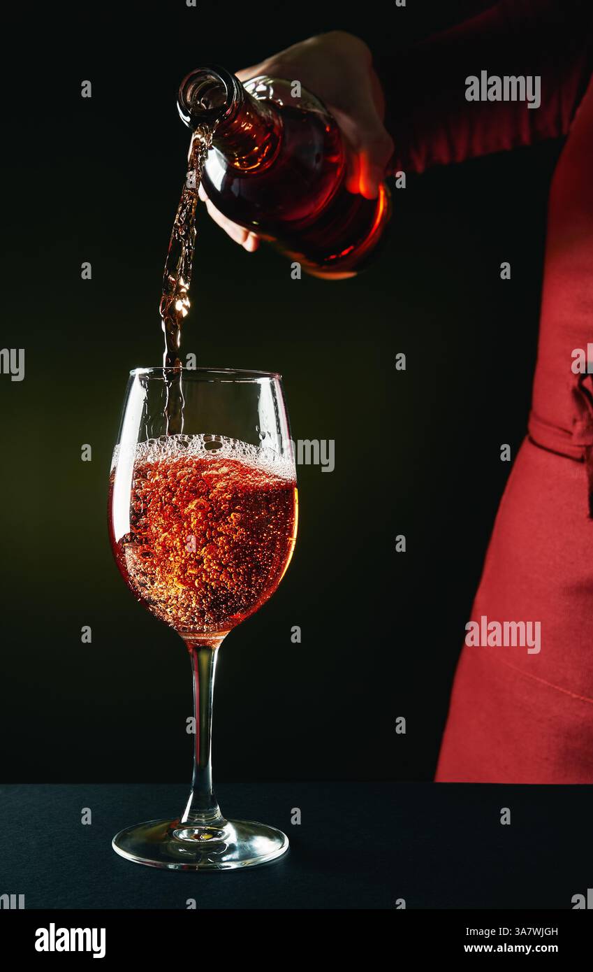 Crop unrecognizable barman in red apron pouring semi sweet fine red wine into wineglass in dark ...