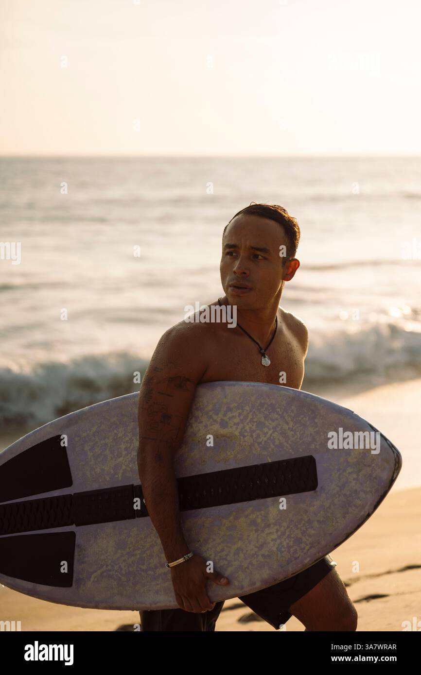 Surfer walking along the beach at sunset, carrying a skimboard under ...