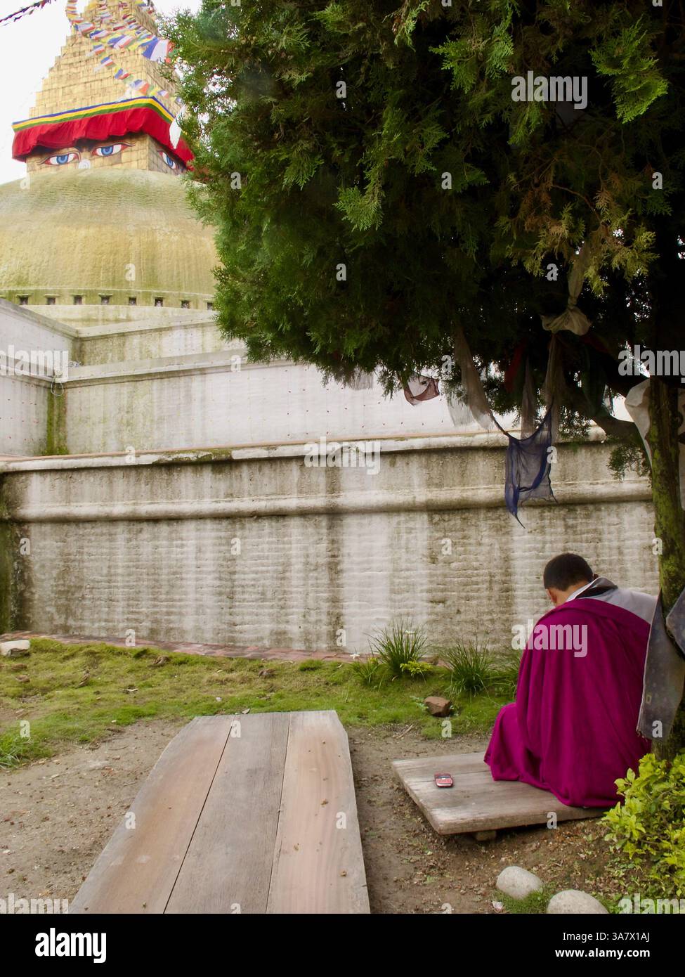 A Buddhist monk sits in silent prayer beneath a tree at the base of Boudhanath Stupa, Kathmandu ...