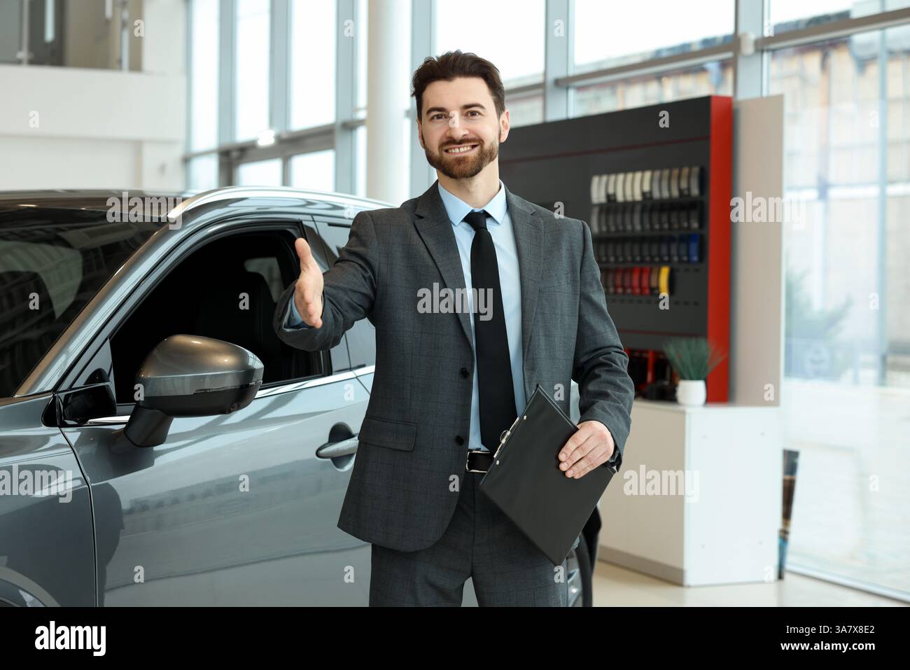 Happy salesman with clipboard offering handshake near new car in salon ...