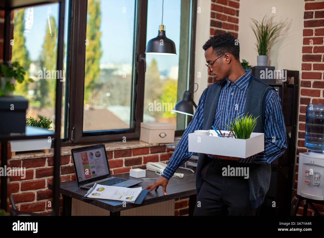 Male employee stands in workspace, holding box containing green plant ...