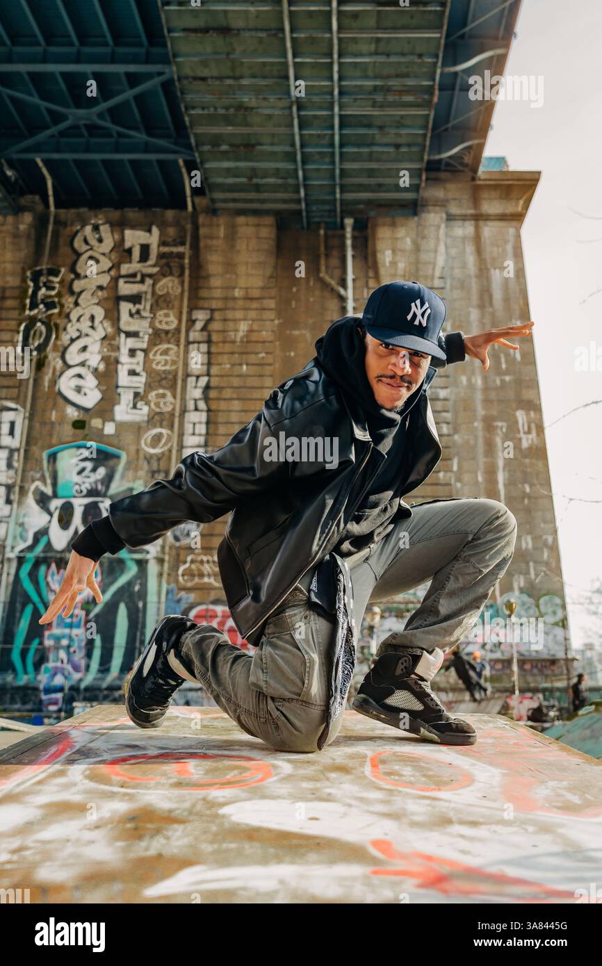Urban dancer kneels in expressive pose under graffiti-covered bridge ...