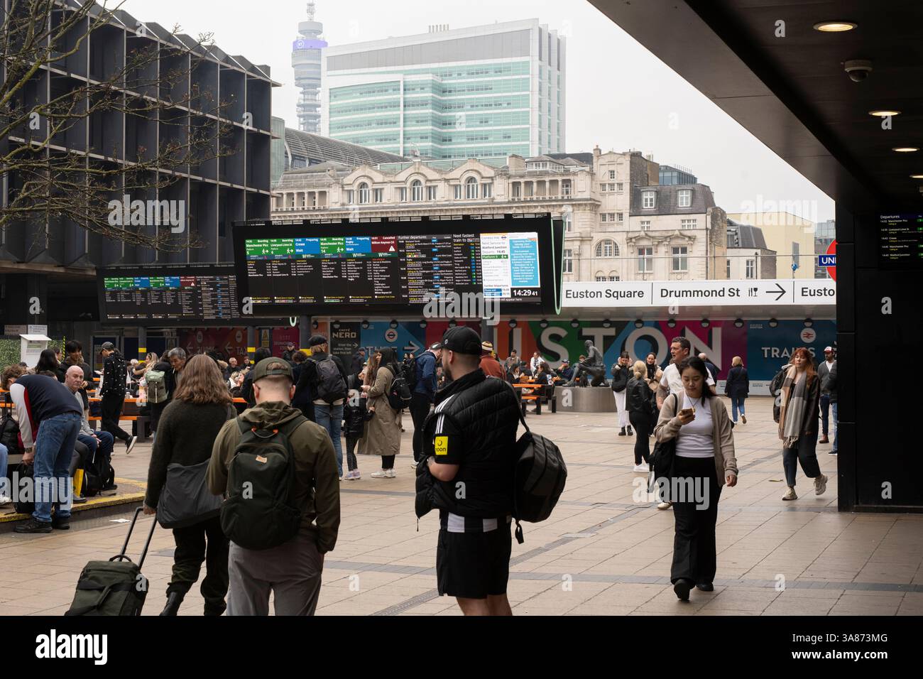 Commuters at Euston Square station in London, UK, await trains. Large digital displays show ...