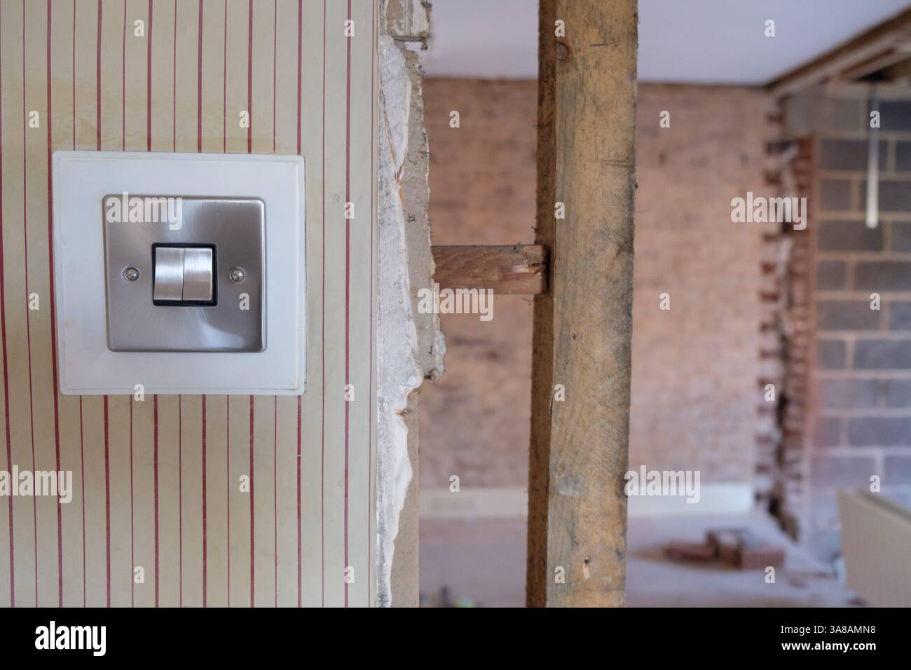 A light switch on striped wallpaper contrasts with a demolished wall section, revealing studs ...