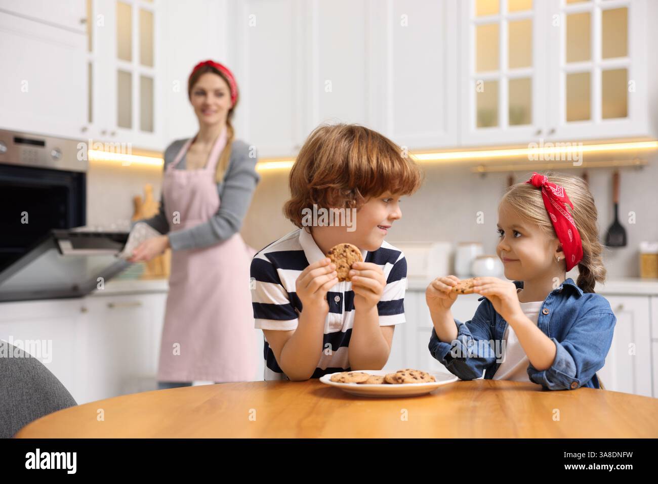 Happy kids eating tasty cookies while their mother baking in kitchen ...