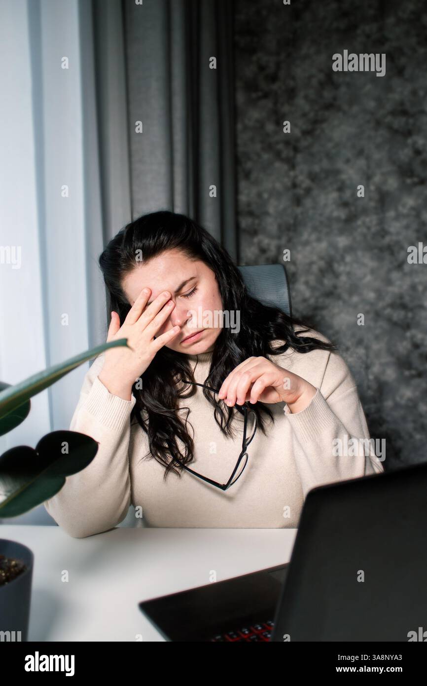 Woman working from home office sitting with laptop rubbing tired eyes ...