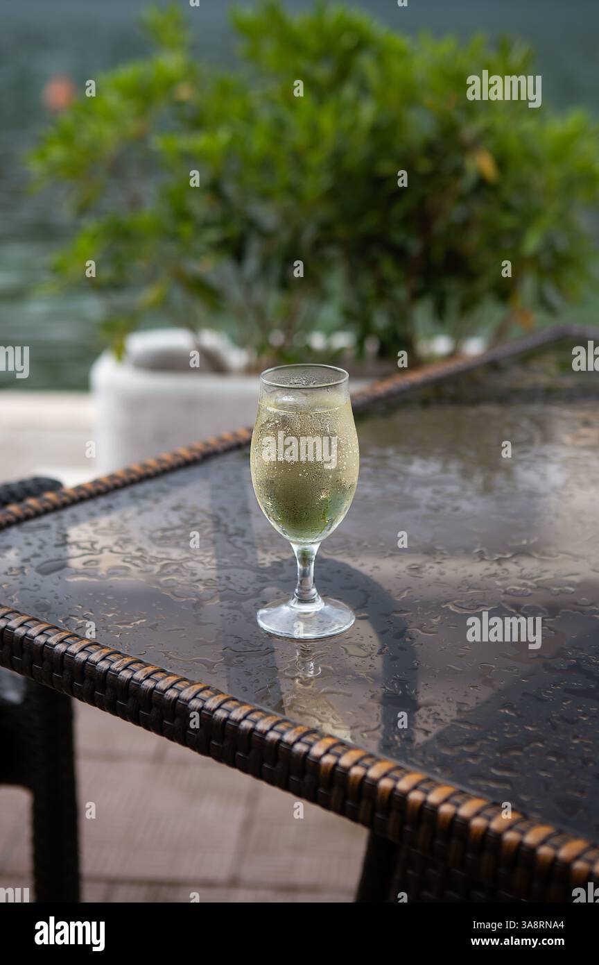 A glass of sparkling white wine sits on a rain-wet outdoor table ...