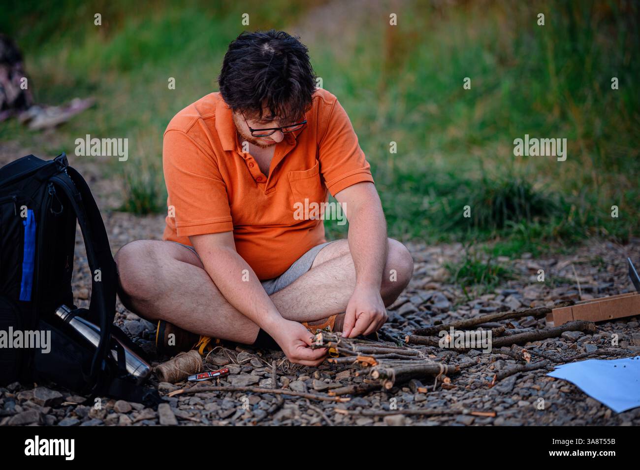 A man sits on rocky ground, tying sticks together to craft a small ...