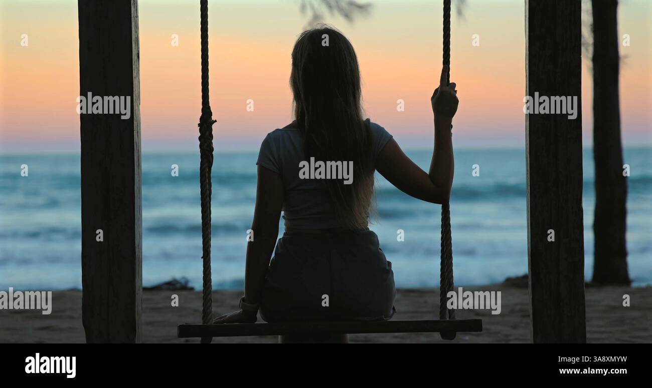 Silhouette of woman on swing facing ocean sunset, peaceful and Stock ...