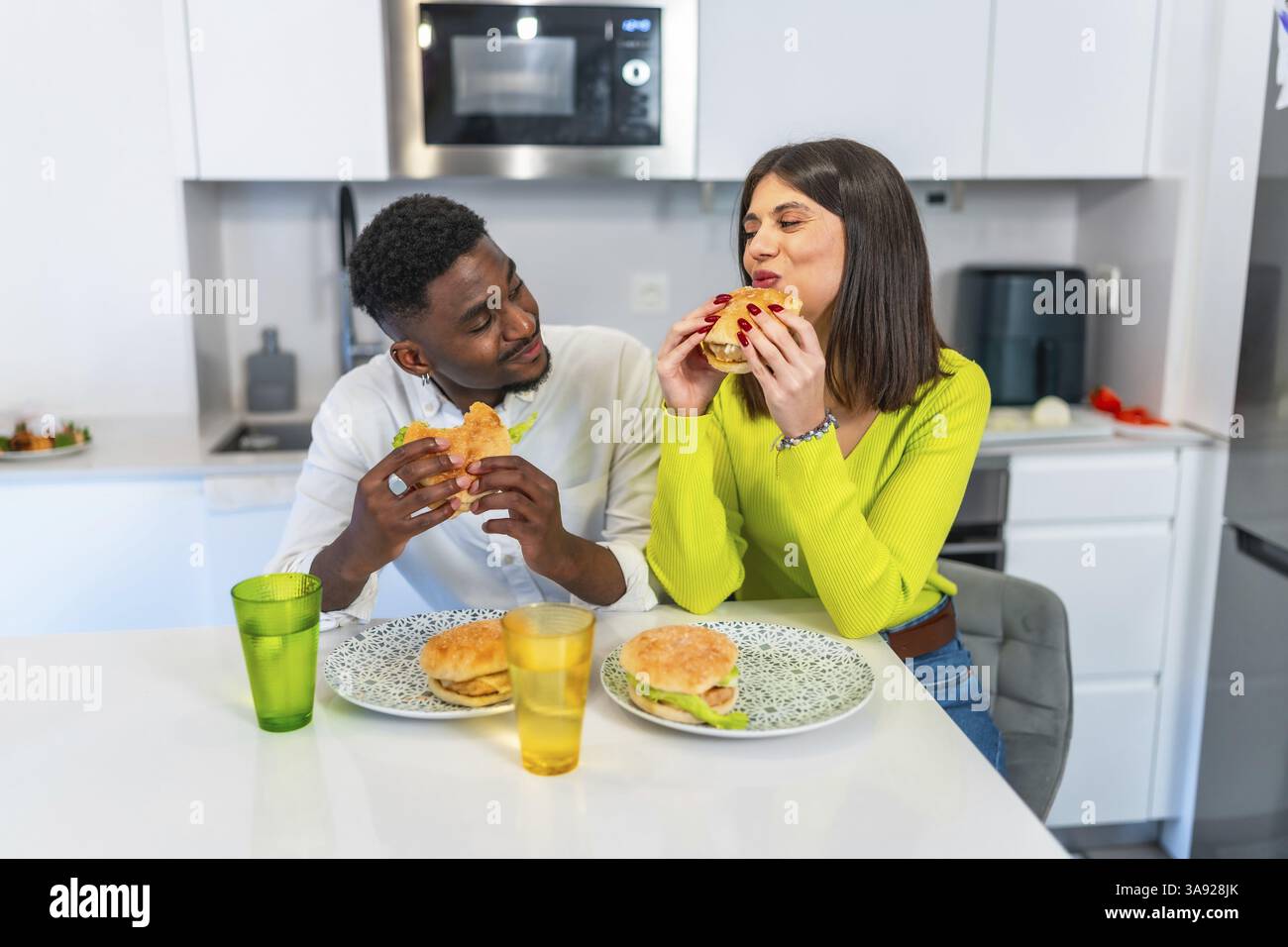 Young multiethnic couple eating burgers in their modern kitchen ...