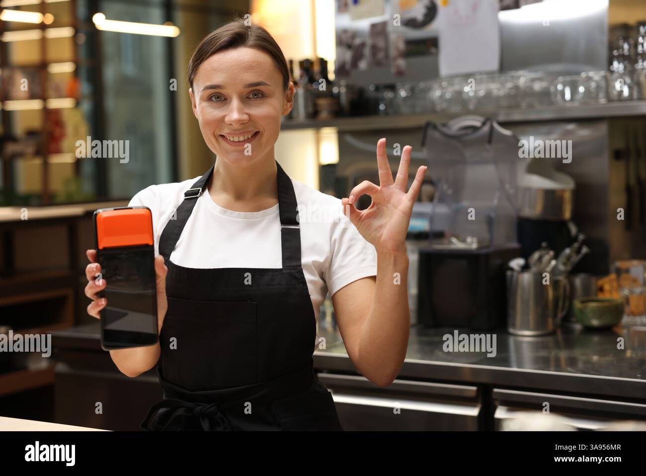 Smiling cafe worker with payment terminal showing OK gesture indoors ...