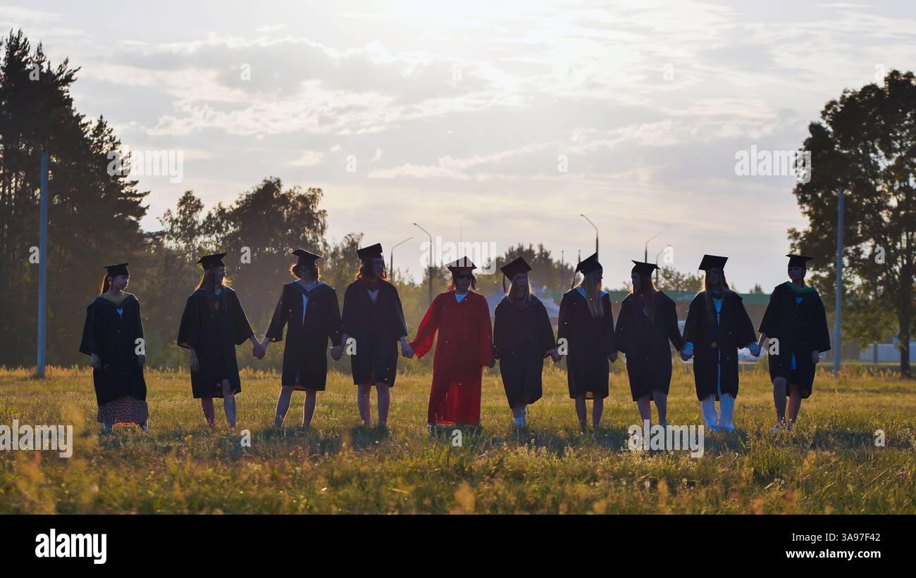 Graduates in academic attire holding hands in a field, celebrating ...