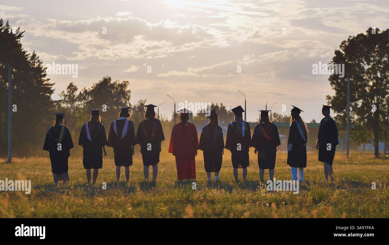 Graduates in academic attire and caps standing in a field at sunset ...