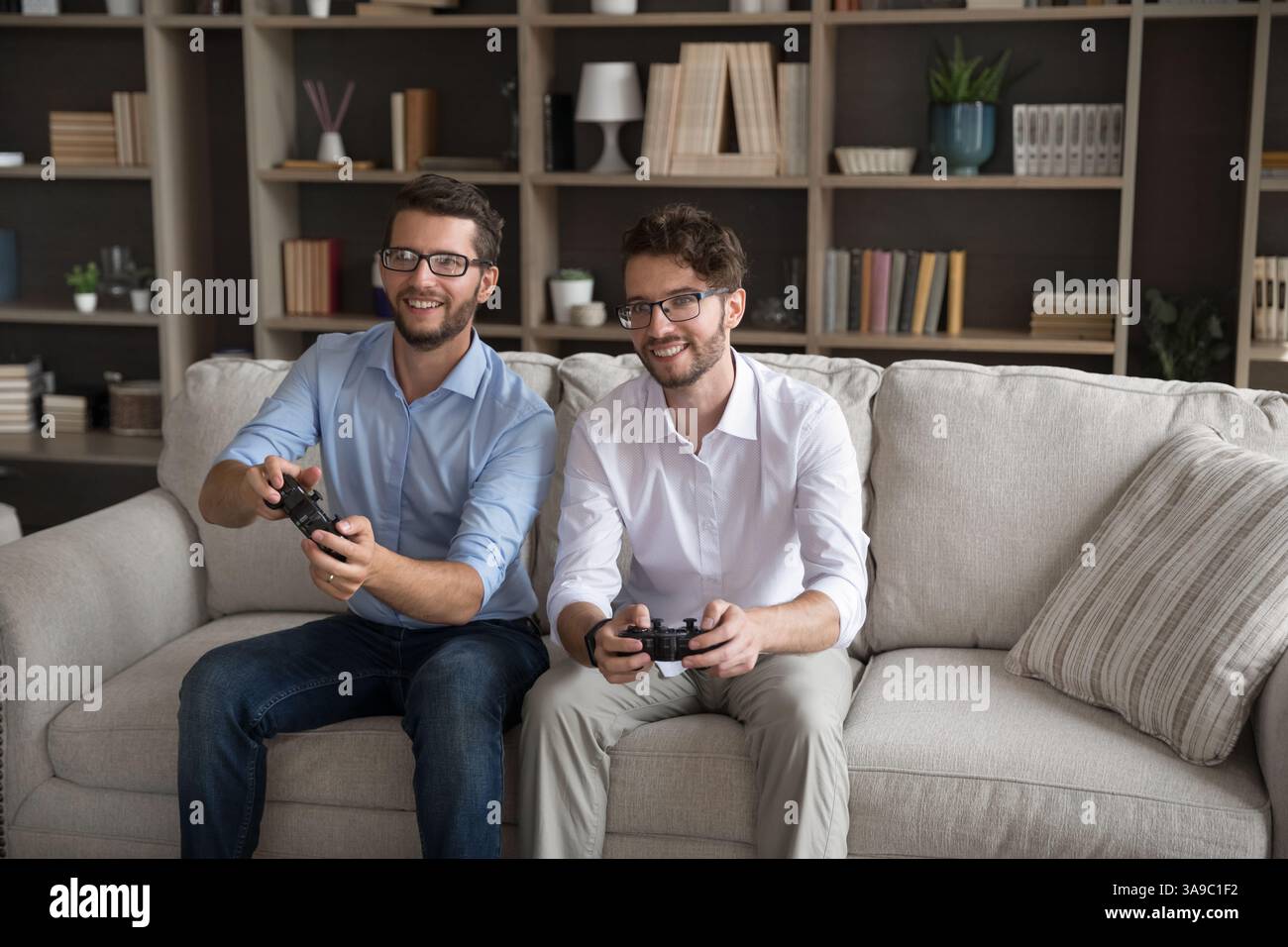 Two millennial twin brothers sitting on sofa playing computer videogame ...