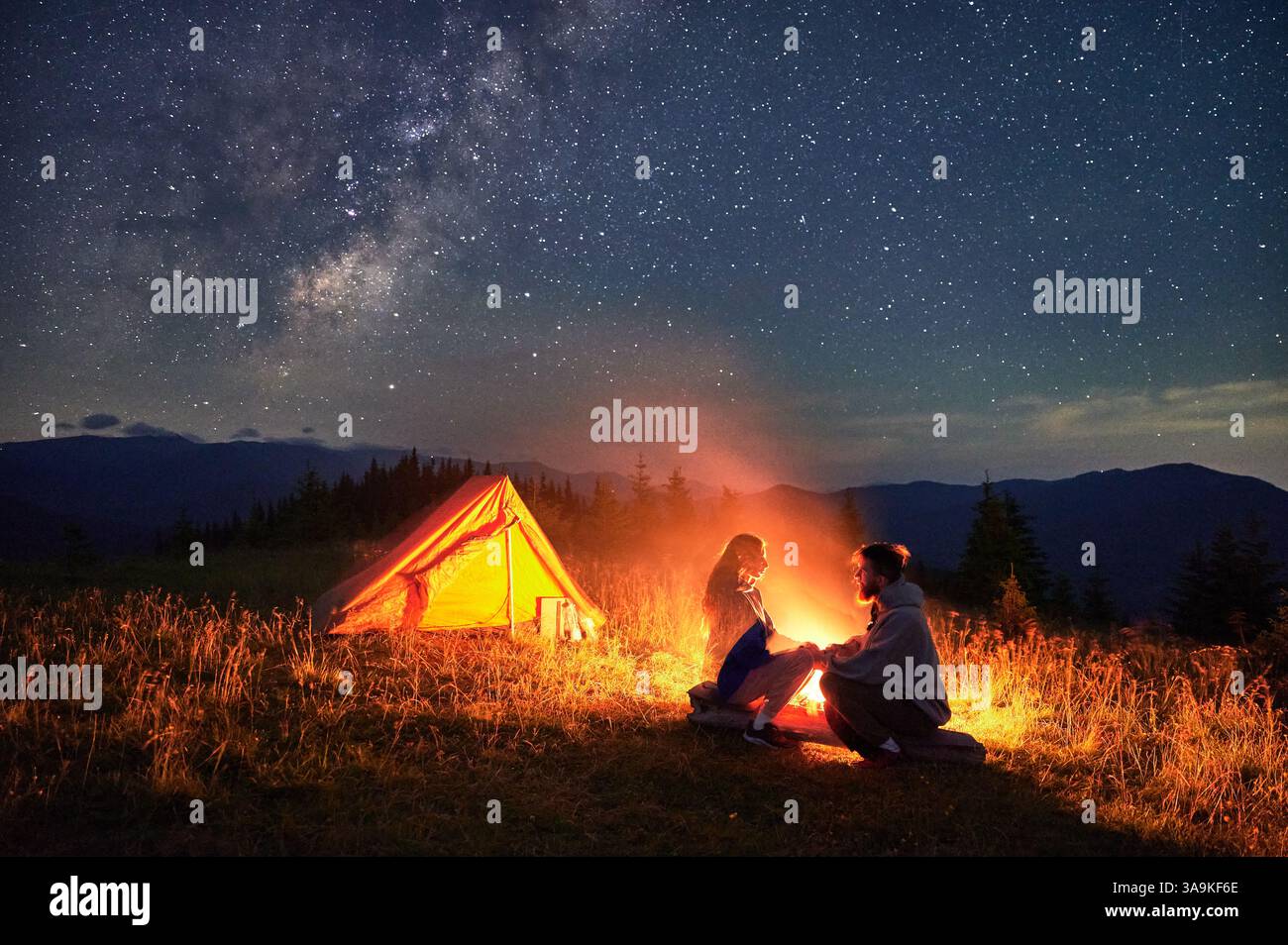 Couple having rest by campfire near their illuminated tent beneath ...