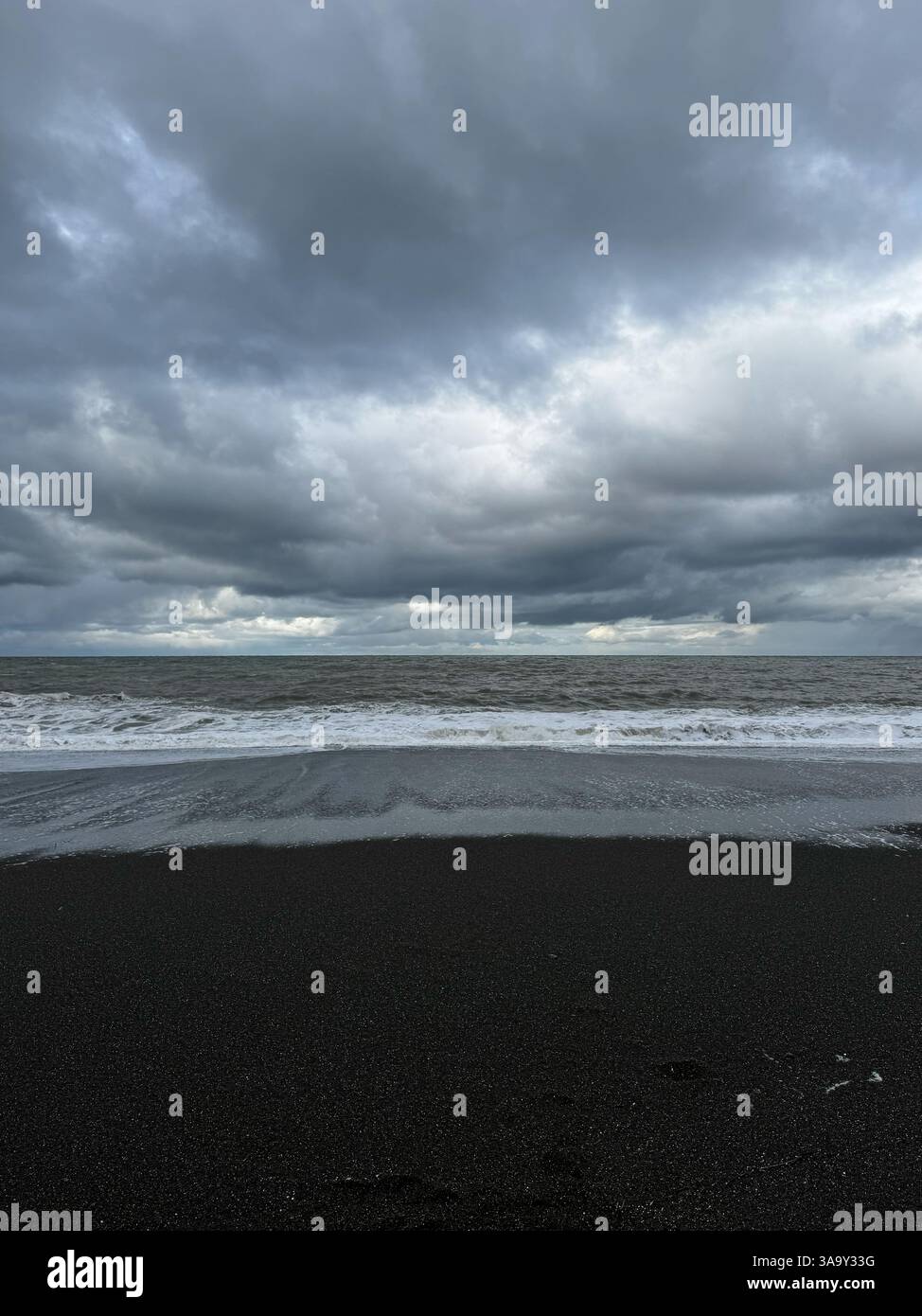 Dark clouds over ocean waves rolling onto a black sand beach, creating ...