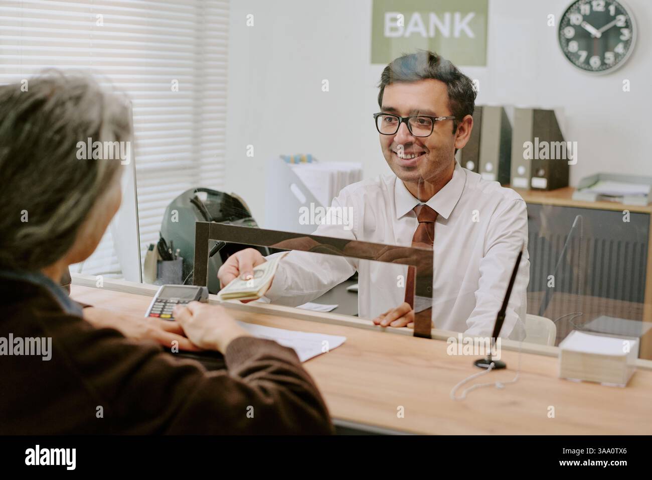 Bank teller wearing glasses and a white shirt smiling while assisting ...