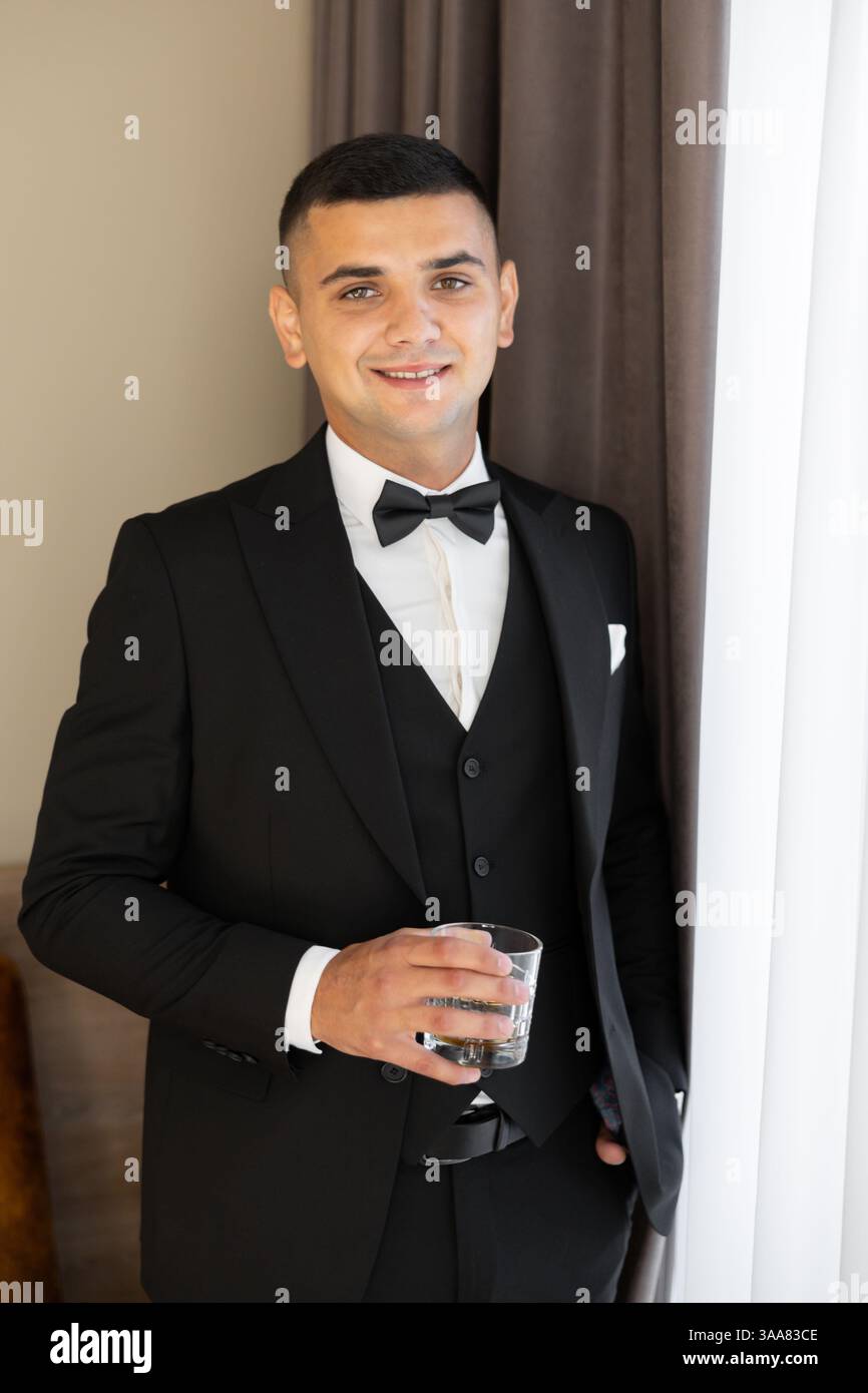 Elegant groom drinking whiskey from a glass while wearing a black suit ...