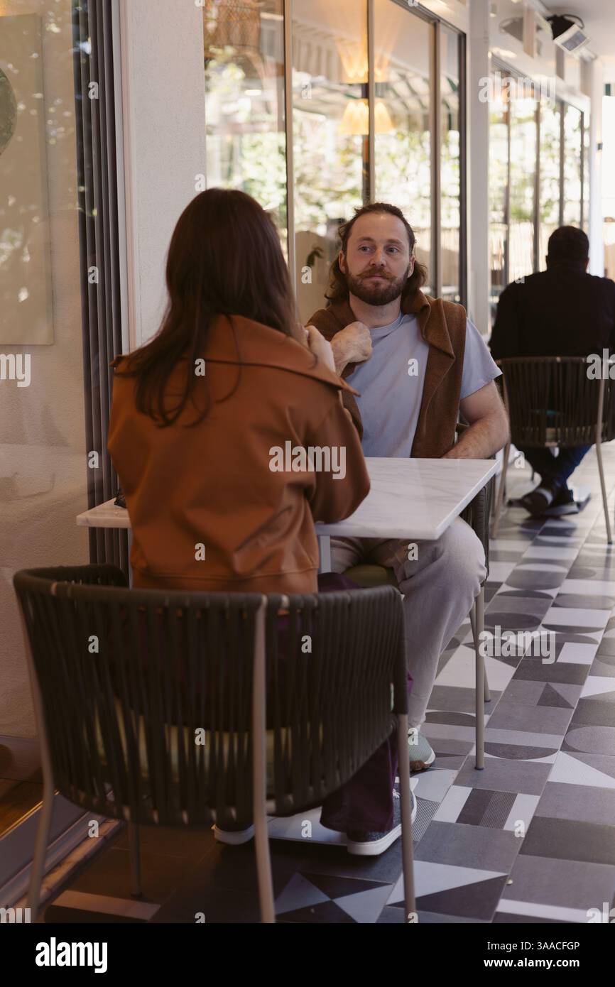 woman and man sit across each other at small table in cozy cafe. They ...